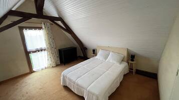 Quarto com teto inclinado, cama com roupa de cama branca, duas mesas de cabeceira com luminárias e uma janela com cortinas.