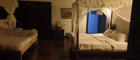 Quarto com duas camas de solteiro, piso de madeira, porta azul e uma pequena mesa com uma cadeira.