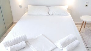 Uma cama bem arrumada com lençóis e almofadas brancas, duas luminárias fixadas na parede e uma pequena mesa redonda com uma cadeira.