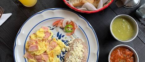 Uma mesa de café da manhã com ovos mexidos, presunto e um croissant em um prato, acompanhados por um copo de suco de laranja, uma cesta de pão e duas pequenas tigelas de acompanhamentos.