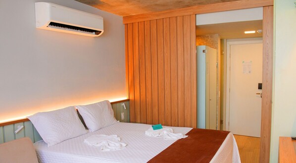 Quarto de hotel com cama, roupa de cama branca, cabeceira de madeira, unidade de ar condicionado e porta que dá para outro quarto.