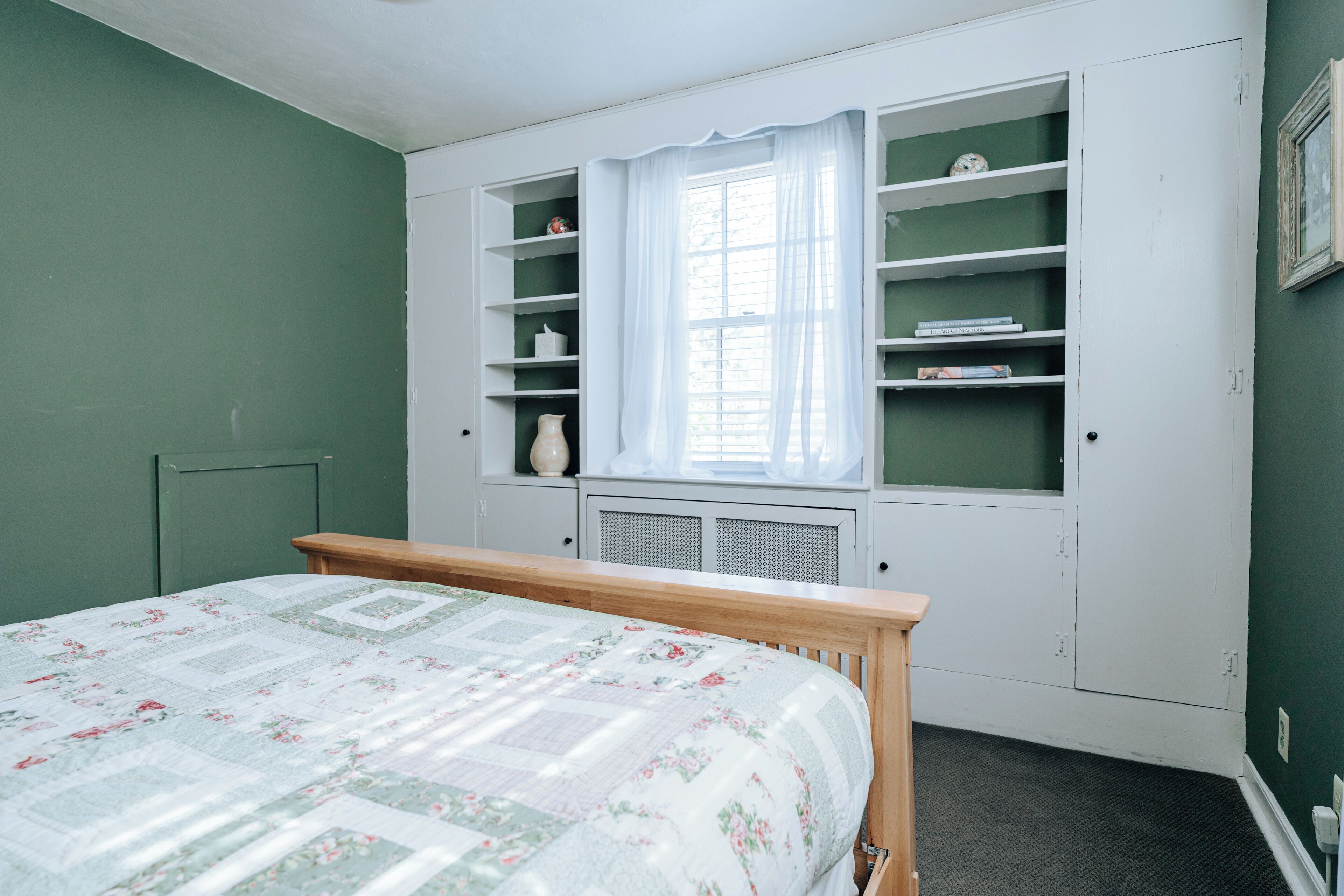 Quarto com cama, janela com cortinas, estante embutida e radiador.