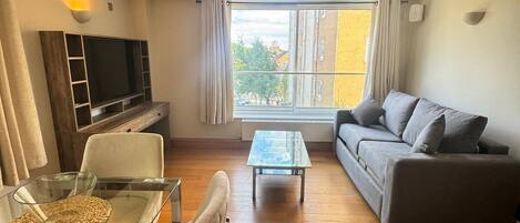 Uma sala de estar com sofá, mesa de centro de vidro, televisão em um móvel e um espaço de jantar com cadeiras e mesa.