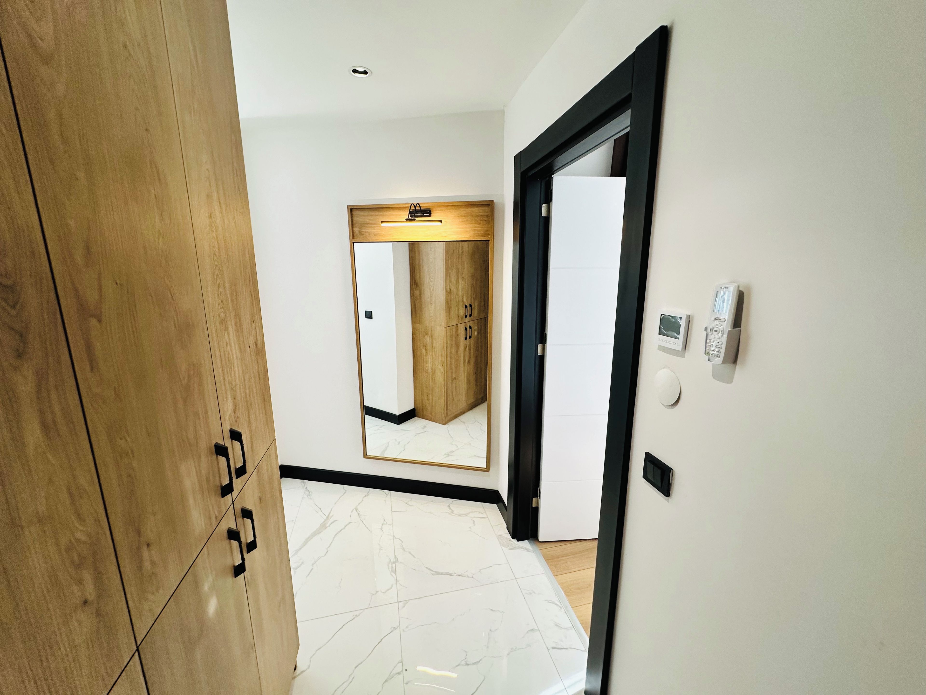 Corredor com porta de madeira, espelho, porta branca e interfone fixado na parede.