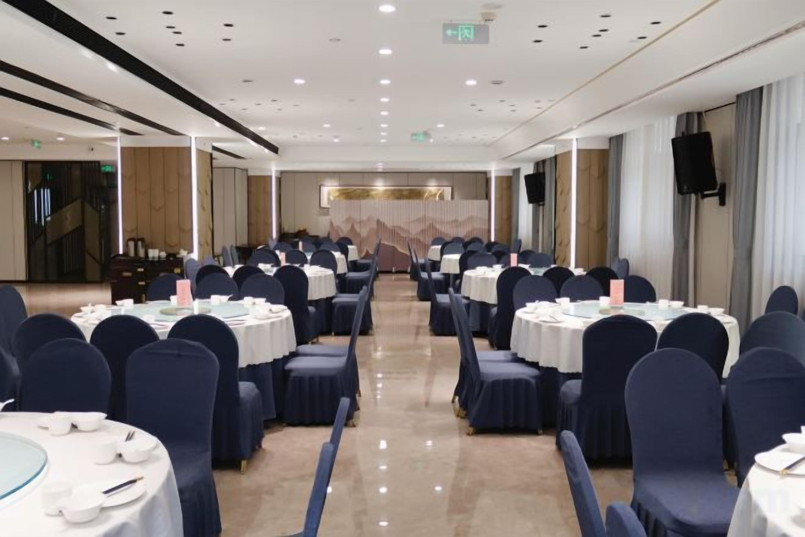 Sala de conferências com fileiras de mesas e cadeiras, palco na frente e escada à esquerda.