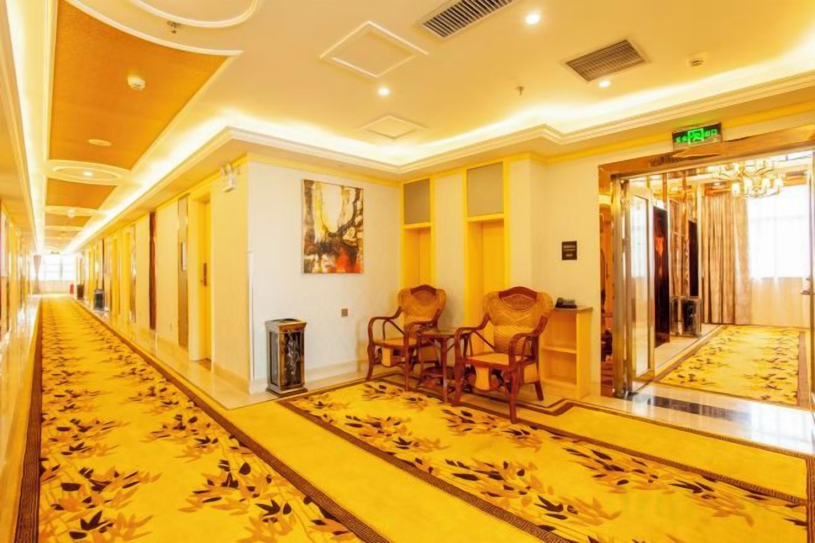 Corredor de hotel com piso carpetado, cadeiras, um cesto de lixo e uma porta que dá para outro quarto.
