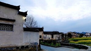 Um pátio tradicional com pavimento de pedra, um edifício com telhado em cerâmica e uma vista distante de um campo.