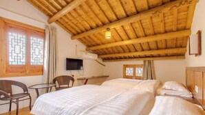 Quarto com teto de madeira, cama com roupa de cama branca, televisão e duas janelas com cortinas.