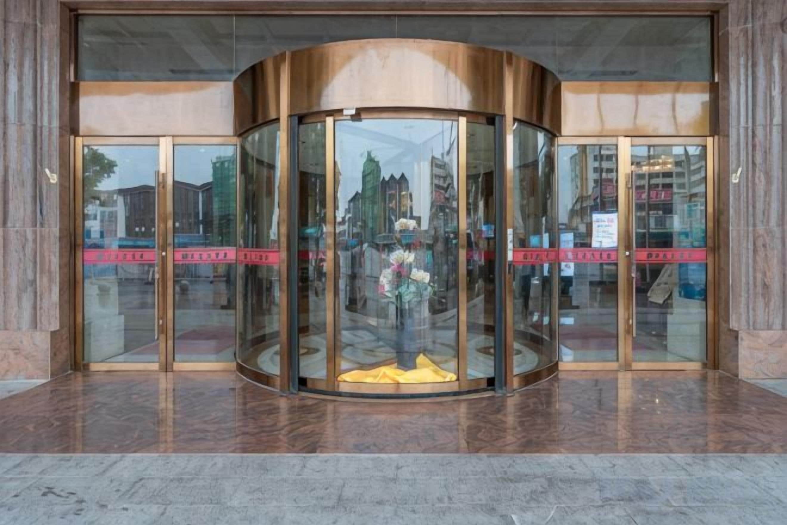 Porta giratória com painéis de vidro, piso de mármore e vista de edifícios ao fundo.