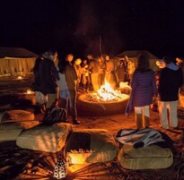 Um grupo de pessoas reunido em volta de uma fogueira à noite, com barracas e sacos de dormir em primeiro plano.