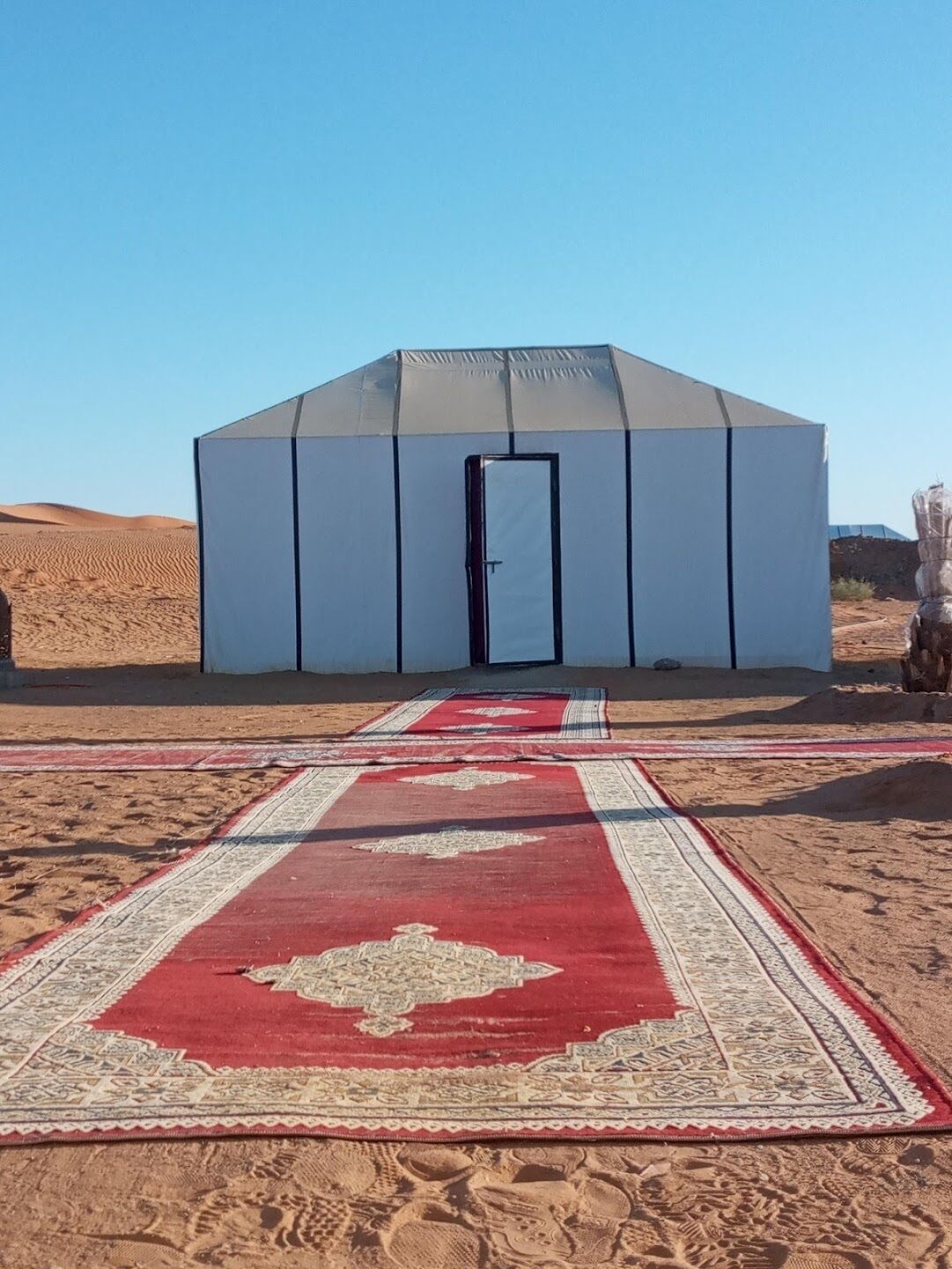 Uma estrutura em forma de tenda com porta, rodeada por dunas de areia e um tapete estampado.