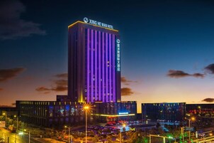 Um hotel alto com linhas verticais iluminadas, um estacionamento e postes de luz.