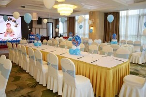 Salão de banquetes com mesas redondas, cadeiras brancas e um palco decorado com balões.