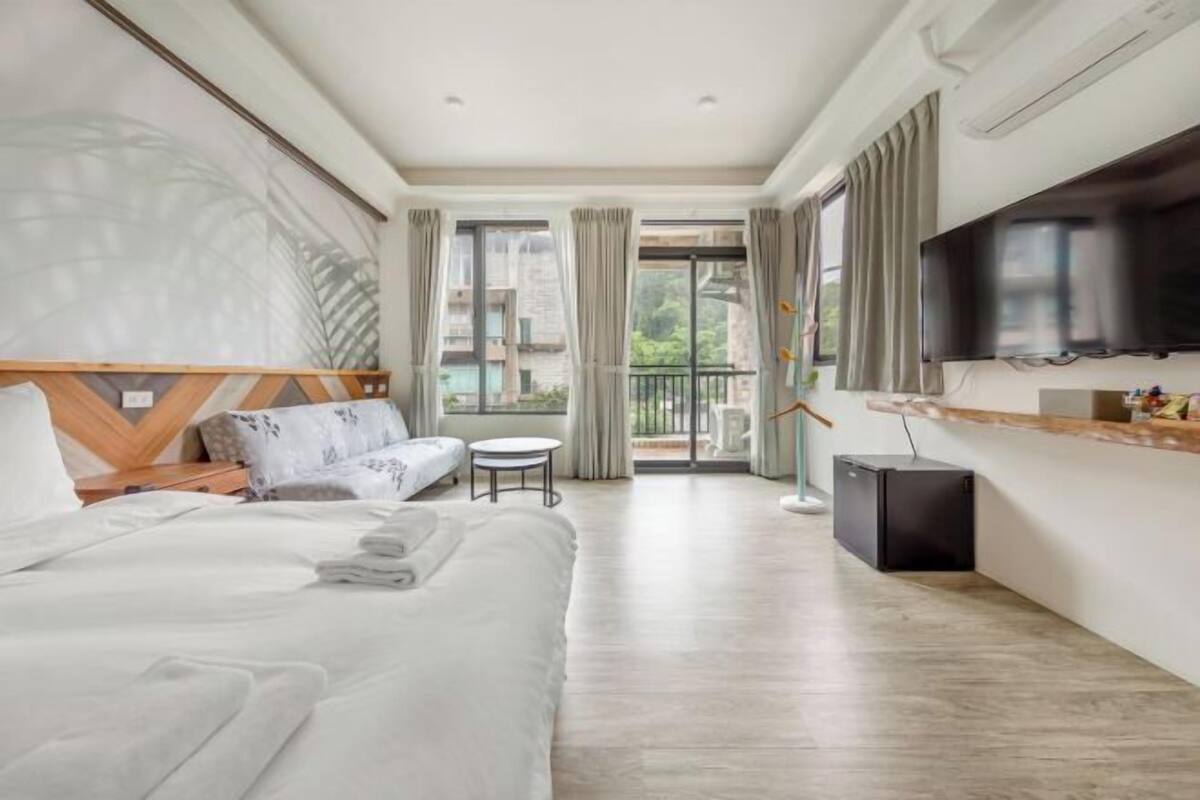 Quarto de hotel com cama, sofá, mesa pequena, televisão e varanda com cortinas.