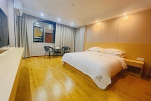 Quarto de hotel com piso de madeira, cama com roupa de cama branca, uma pequena escrivaninha, duas cadeiras e uma janela com cortinas.