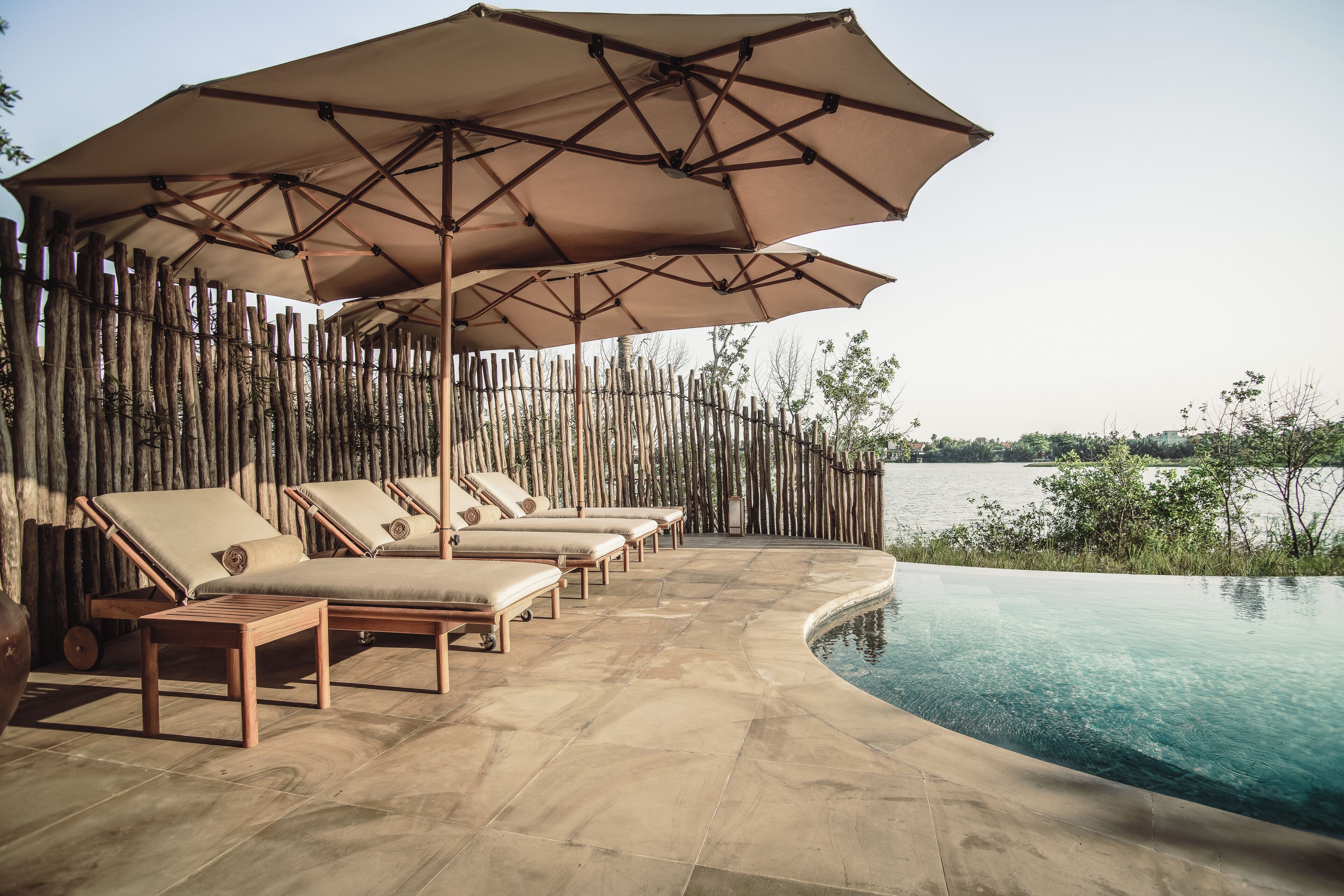 Uma área à beira da piscina com espreguiçadeiras e guarda-sóis, um cercado de bambu e vista para um corpo d'água.