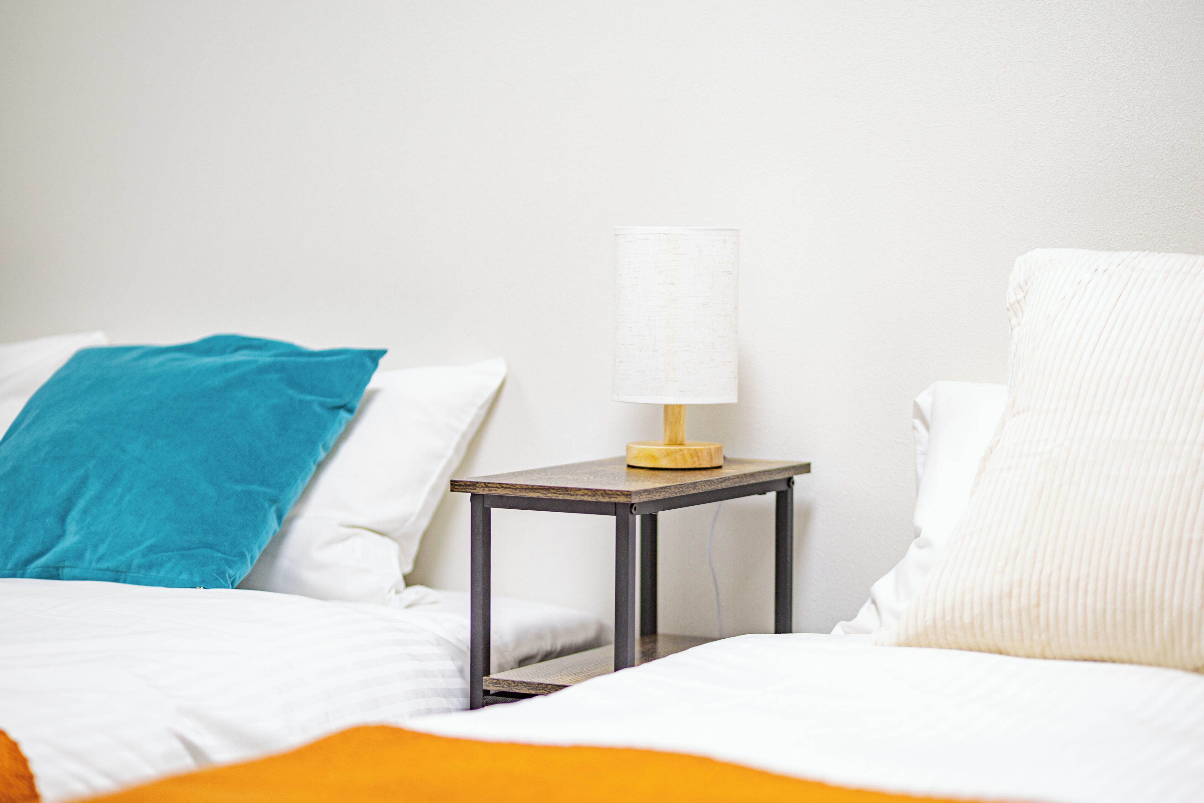 Uma cama com roupa de cama branca, um travesseiro azul-turquesa e um travesseiro branco. Há uma pequena mesa com um abajur entre os travesseiros.