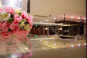 Um hall de hotel com uma superfície refletora, um arranjo floral e um balcão de recepção.