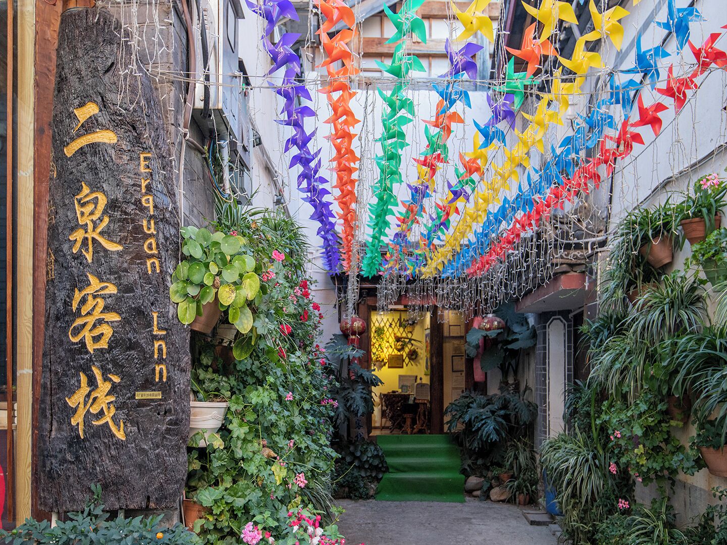 Um beco estreito com moinhos de vento coloridos pendurados acima, uma placa de madeira com caracteres chineses e vegetação exuberante em ambos os lados.