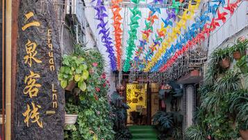 Um beco estreito com moinhos de vento coloridos pendurados acima, uma placa de madeira com caracteres chineses e vegetação exuberante em ambos os lados.