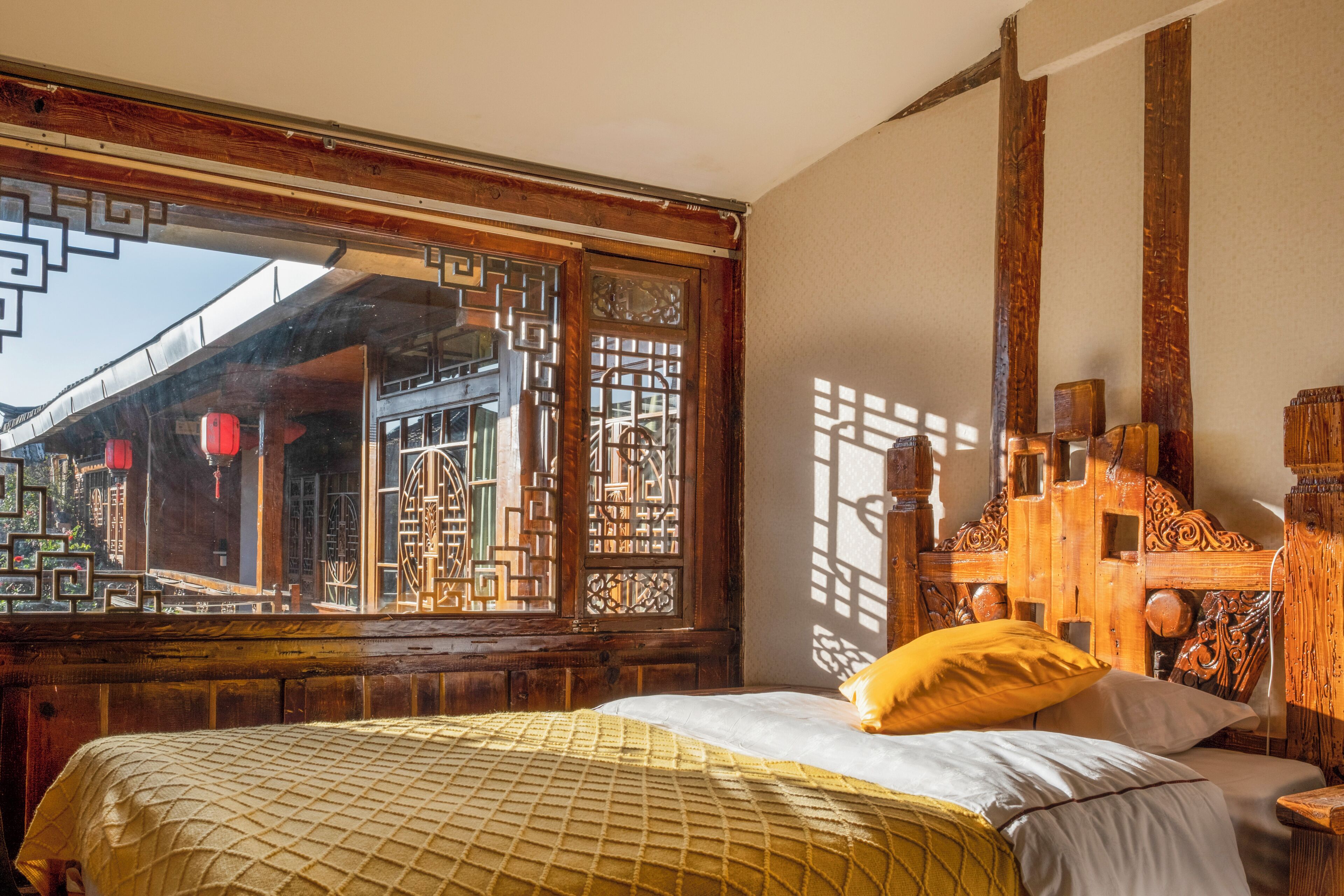 Uma cama de madeira tradicional com colcha amarela, cabeceira de madeira com esculturas detalhadas e janela com grade de madeira geométrica.