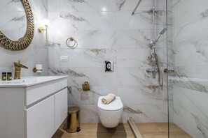 Um banheiro moderno com piso de mármore, vaso sanitário branco, lixeira dourada, box de vidro e um espelho grande com moldura dourada.