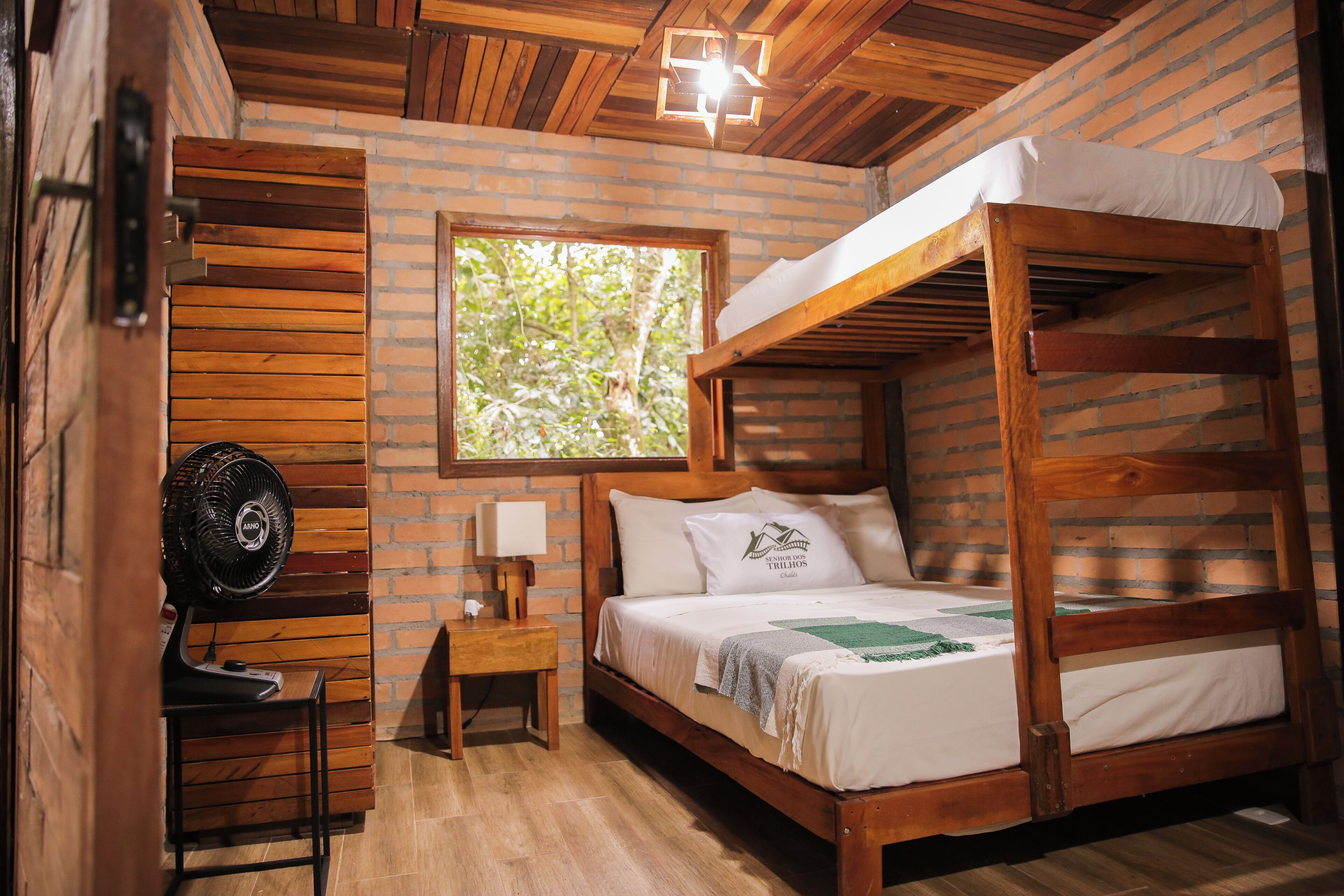 Quarto com beliche de madeira, um ventilador, uma mesinha e janela com vista para árvores.