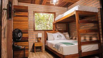 Quarto com beliche de madeira, um ventilador, uma mesinha e janela com vista para árvores.