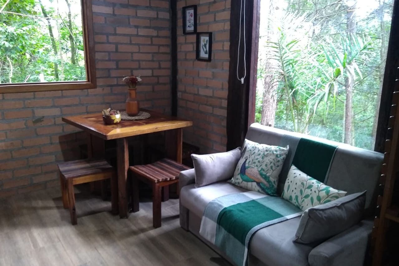 Um quarto aconchegante com parede de tijolos, uma mesa de madeira com um vaso, um sofá cinza com almofadas estampadas e uma janela com vista para o verde.