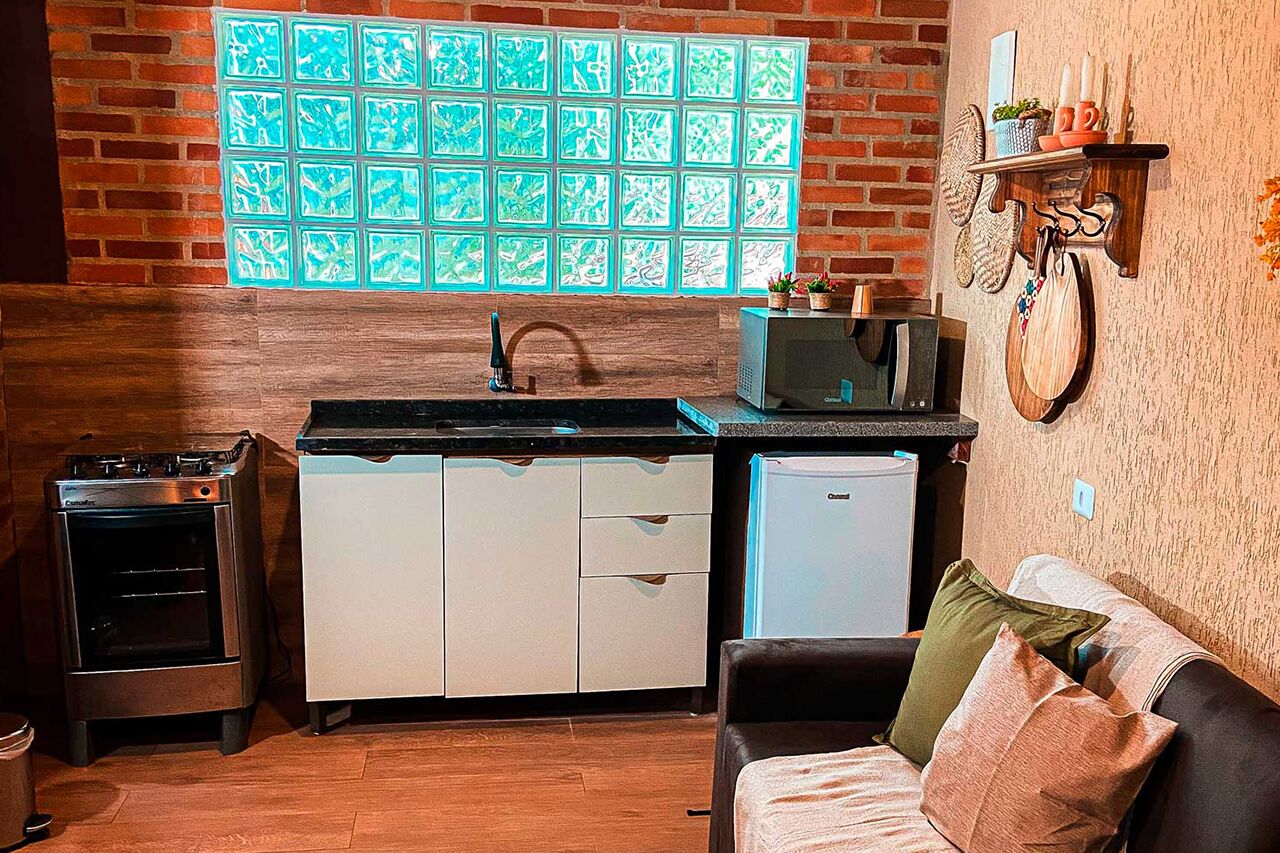Uma cozinha compacta com parede de tijolos, janela com padrão quadriculado, micro-ondas e geladeira pequena.