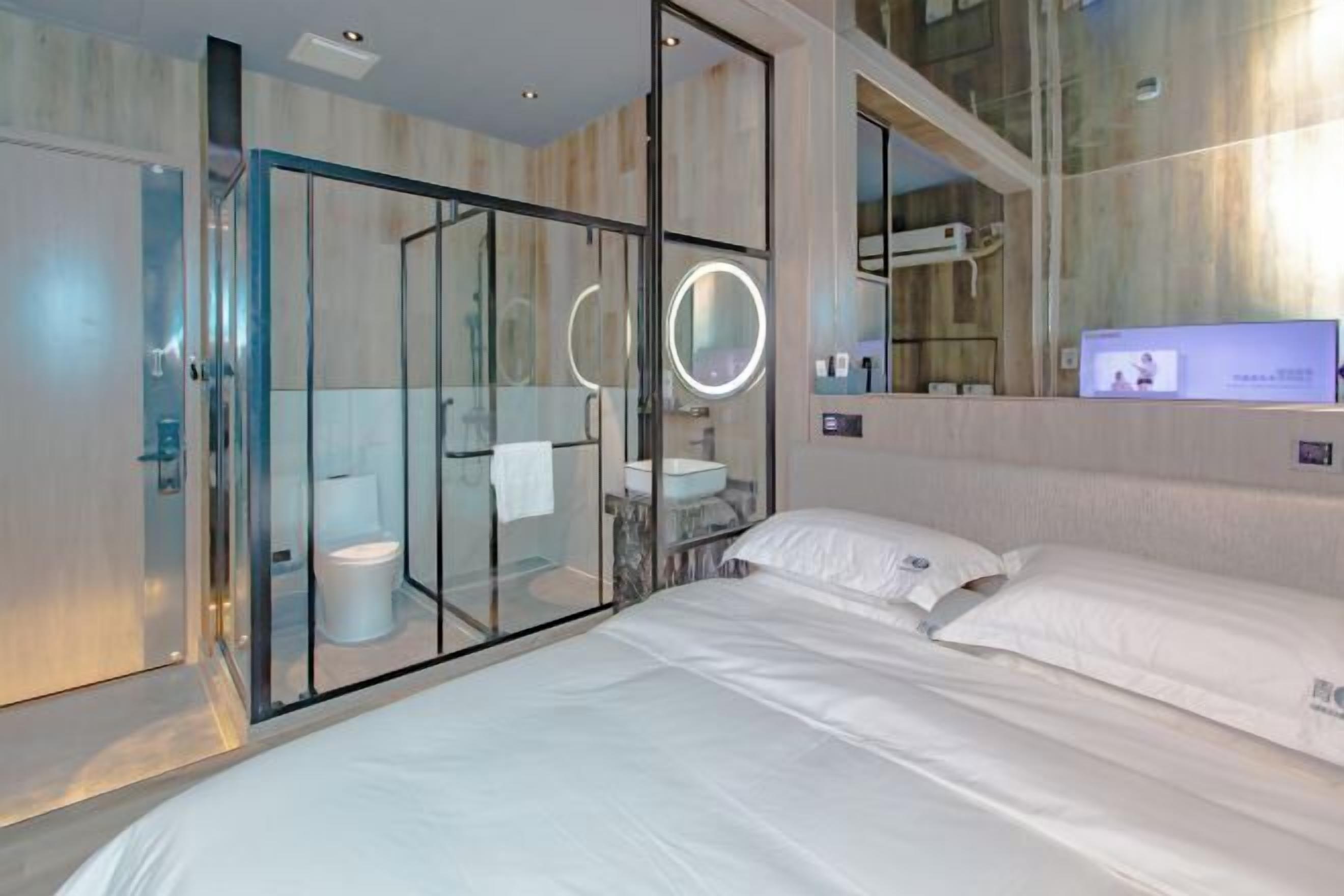 Quarto de hotel moderno com chuveiro em gabinete de vidro, cama com roupa de cama branca e TV embutida na parede.