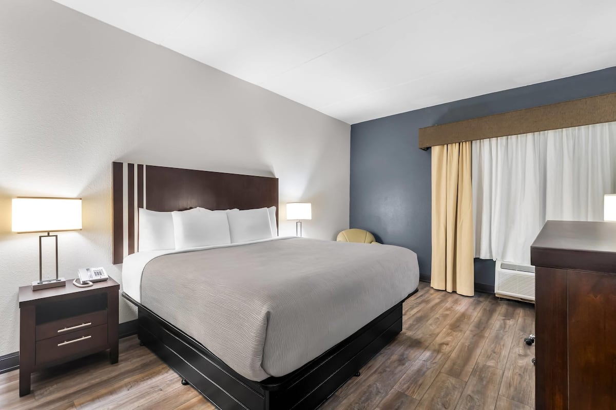 Quarto de hotel com cama, mesas de cabeceira, luminária e janela com cortinas.