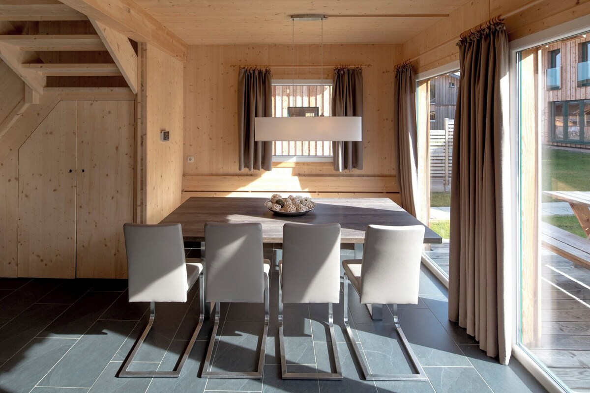 Uma área de refeições com mesa e cadeiras de madeira, uma tigela com objetos decorativos e janelas amplas com cortinas.