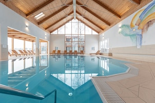 Piscina interna com vigas de madeira, paredes de vidro transparente e vista para o exterior.
