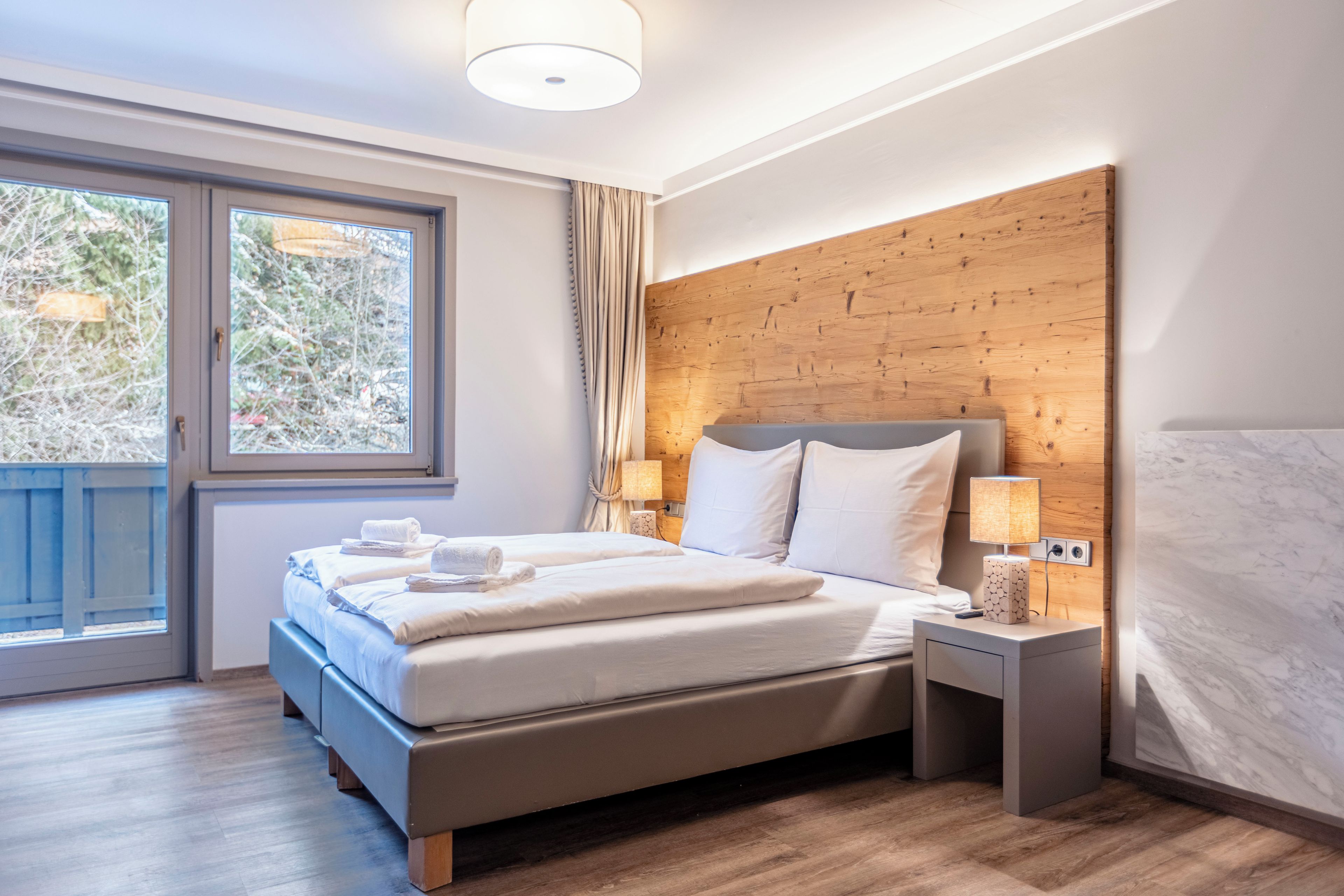 Um quarto moderno com cabeceira de madeira, uma cama grande com roupa de cama branca, duas mesas de cabeceira com luminárias e vista para árvores pela janela.