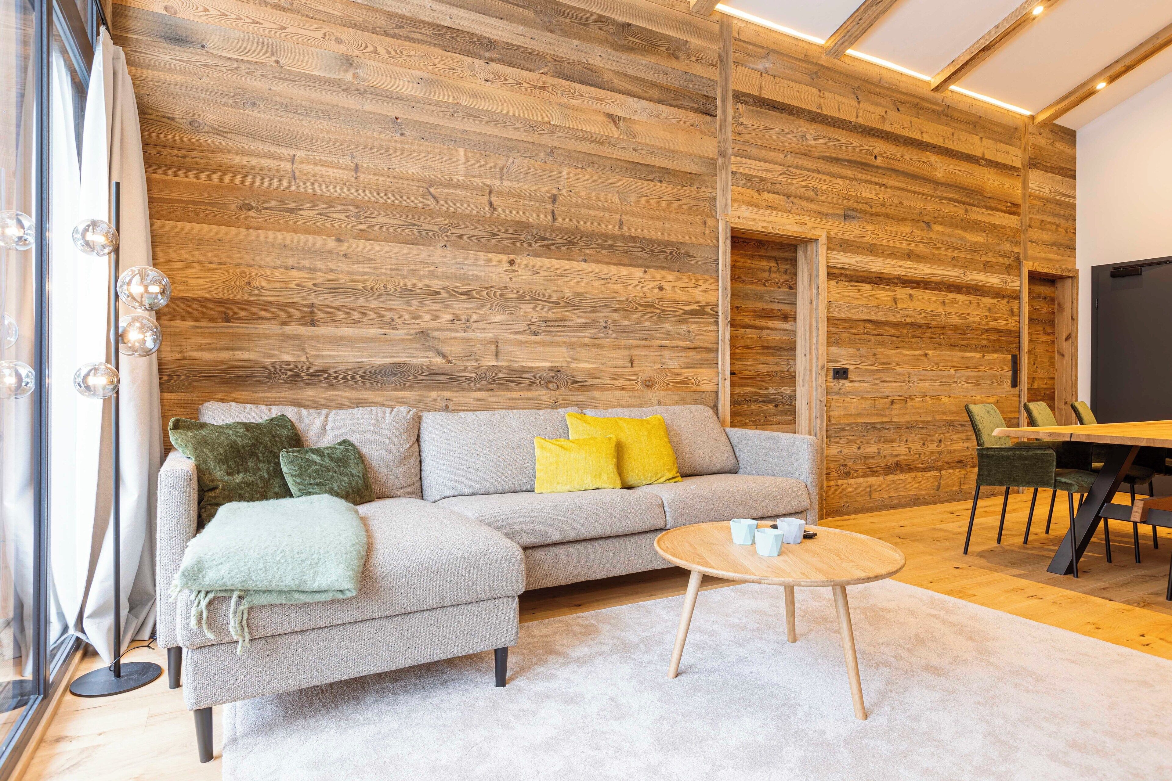 Uma sala de estar moderna com parede de madeira, sofá cinza com almofadas amarelas e verdes, mesa de centro redonda e um espaço de jantar com mesa preta e cadeiras verdes.