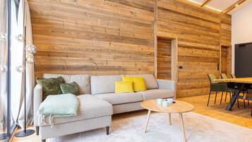 Uma sala de estar moderna com parede de madeira, sofá cinza com almofadas amarelas e verdes, mesa de centro redonda e um espaço de jantar com mesa preta e cadeiras verdes.