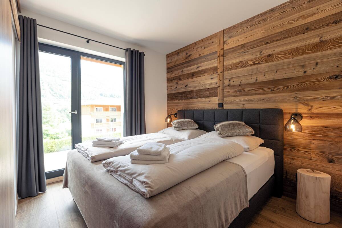 Quarto de hotel moderno com uma cama grande, painel de parede de madeira e vista para o exterior.