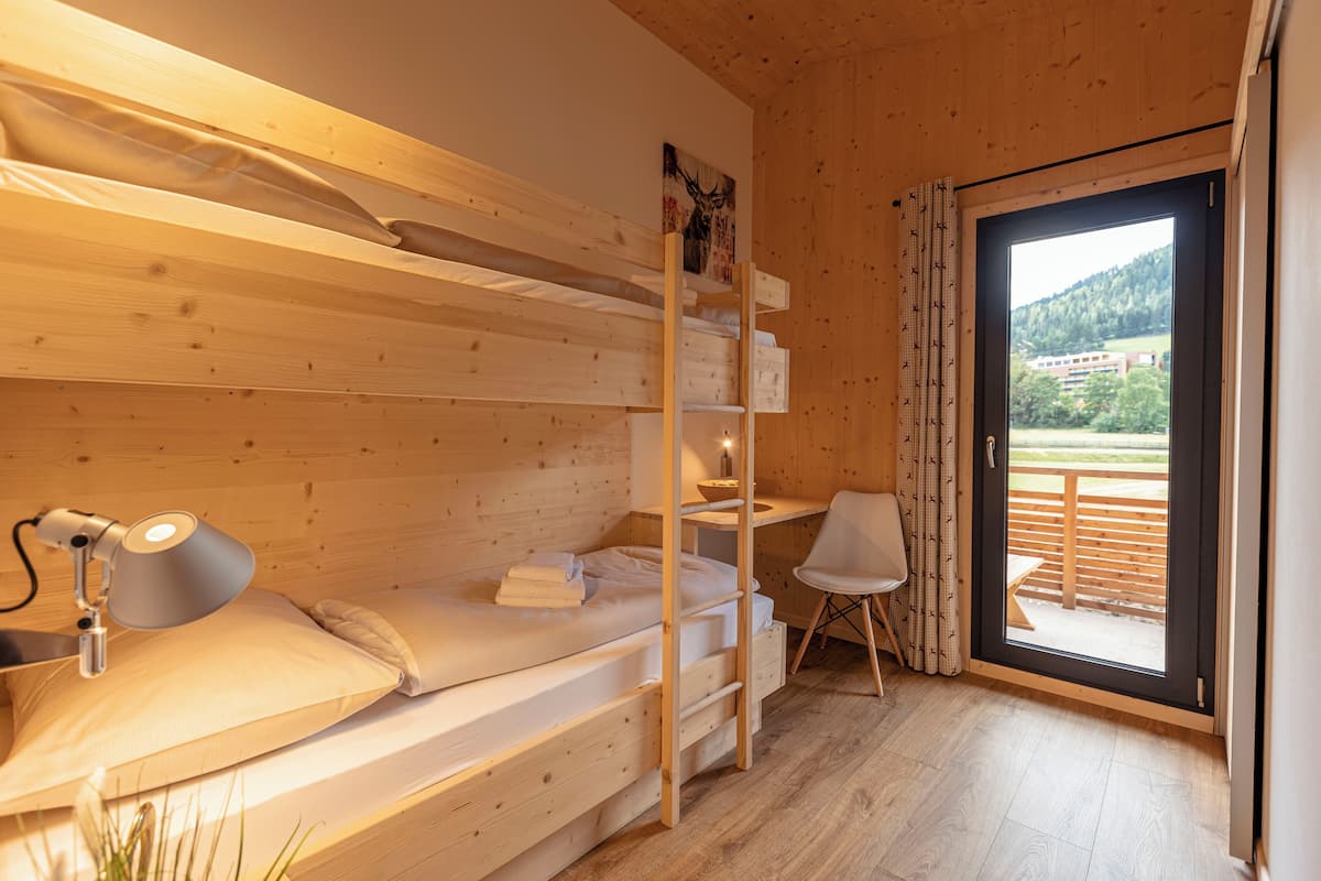 Quarto de cabana de madeira com beliche, escrivaninha com luminária, cadeira e vista para uma floresta através de uma porta de vidro.