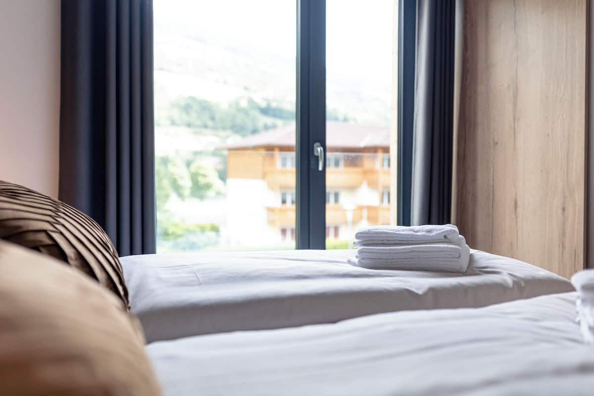 Quarto de hotel com cama, janela com cortinas e vista para um edifício ao fundo.