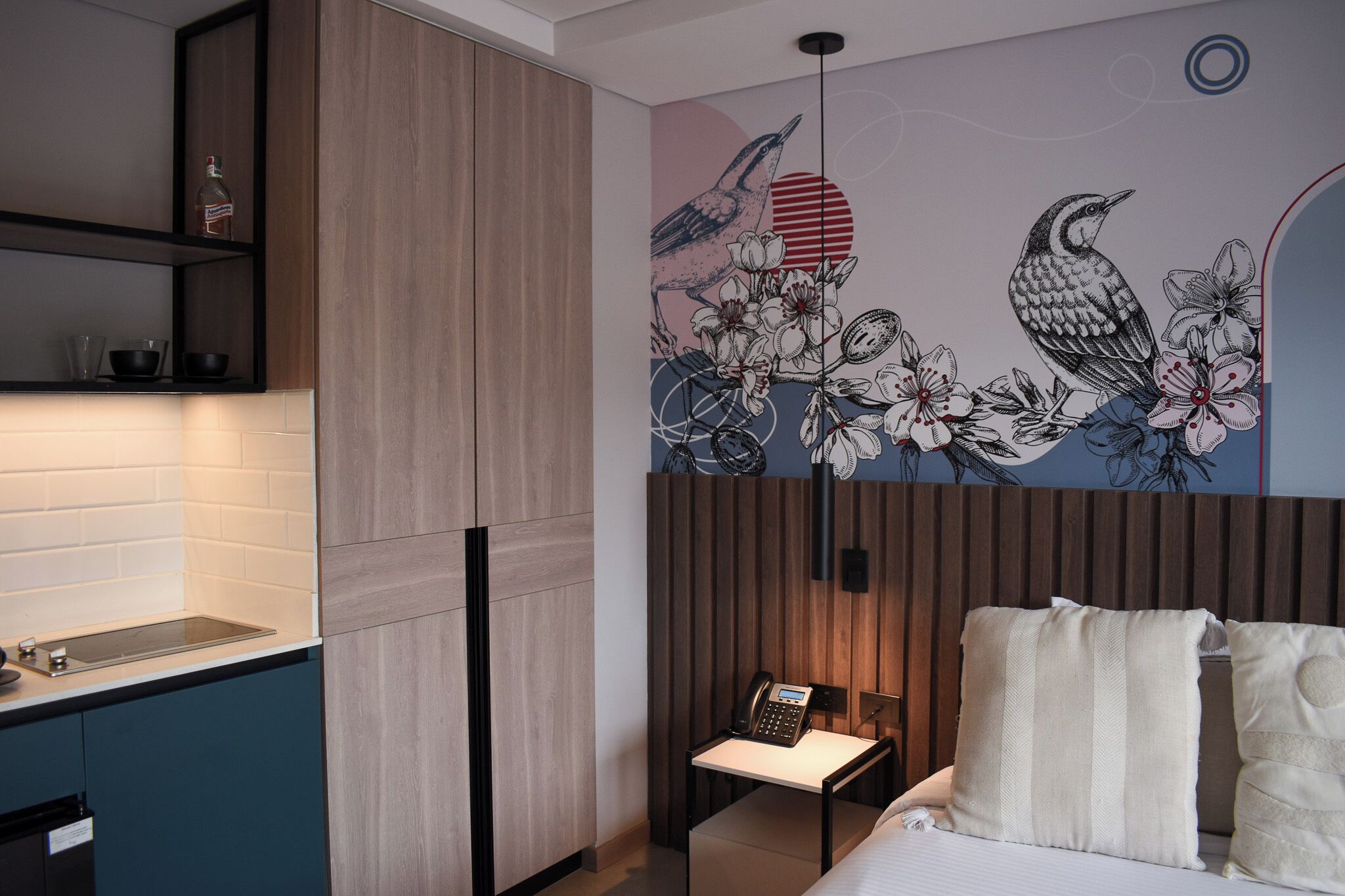 Quarto de hotel moderno com toucador de madeira, cama com roupa de cama branca, mesinha de cabeceira com telefone e um mural na parede com pássaros e flores.