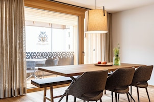 Uma sala de jantar com uma mesa de madeira, quatro cadeiras, um lustre e vista para o exterior por grandes janelas.