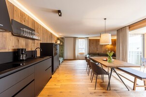 Cozinha moderna com bancada de madeira, cafeteira e forno embutido. Há uma área de refeições com mesa e cadeiras de madeira, e um banco.