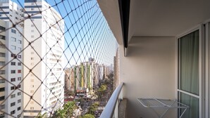 Varanda com rede de proteção metálica, janela com cortina e vista para paisagem urbana com edifícios.