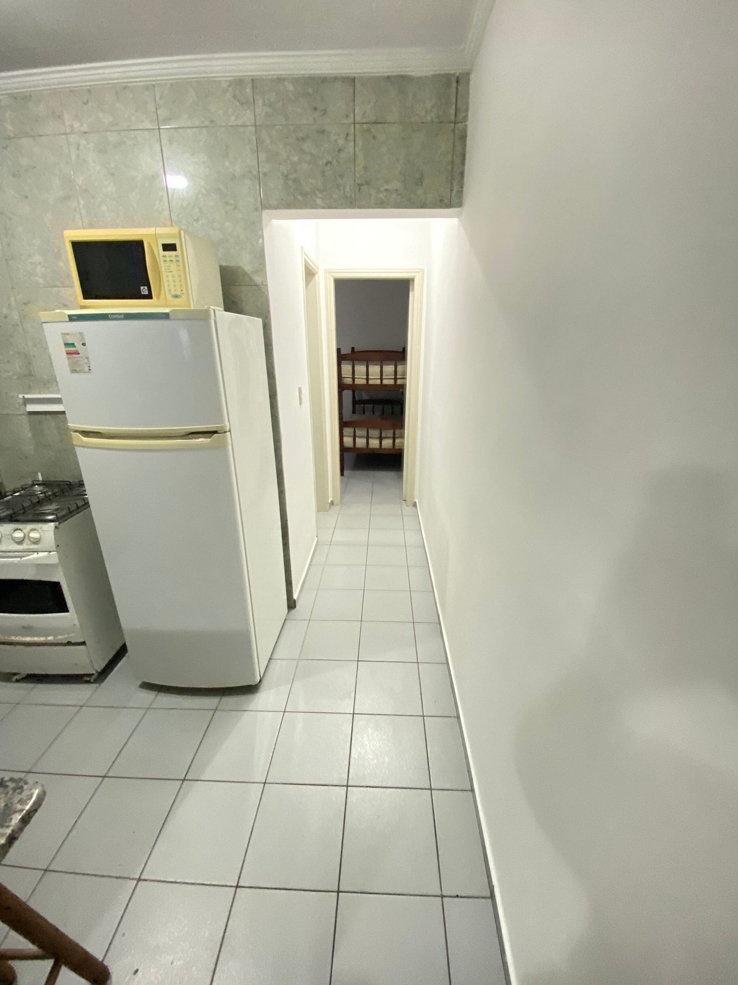Cozinha pequena com geladeira, fogão e micro-ondas. Uma porta dá acesso a um corredor com uma cama.