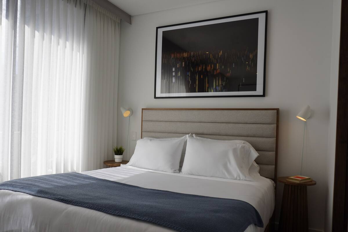 Quarto de hotel moderno com uma cama grande, uma pintura de paisagem urbana sobre a cama, uma mesa de cabeceira com abajur e uma janela com cortinas leves.
