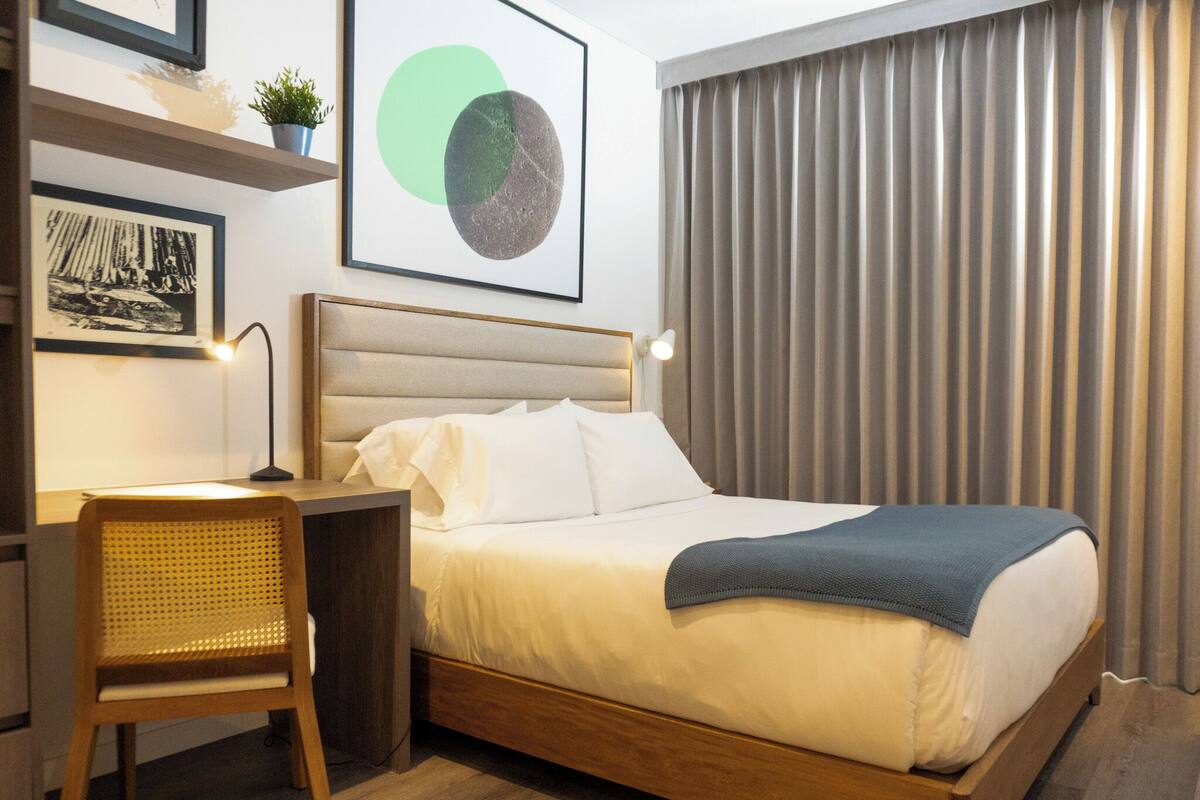 Quarto de hotel bem arrumado, com cama, escrivaninha, cadeira e uma janela grande com cortinas.