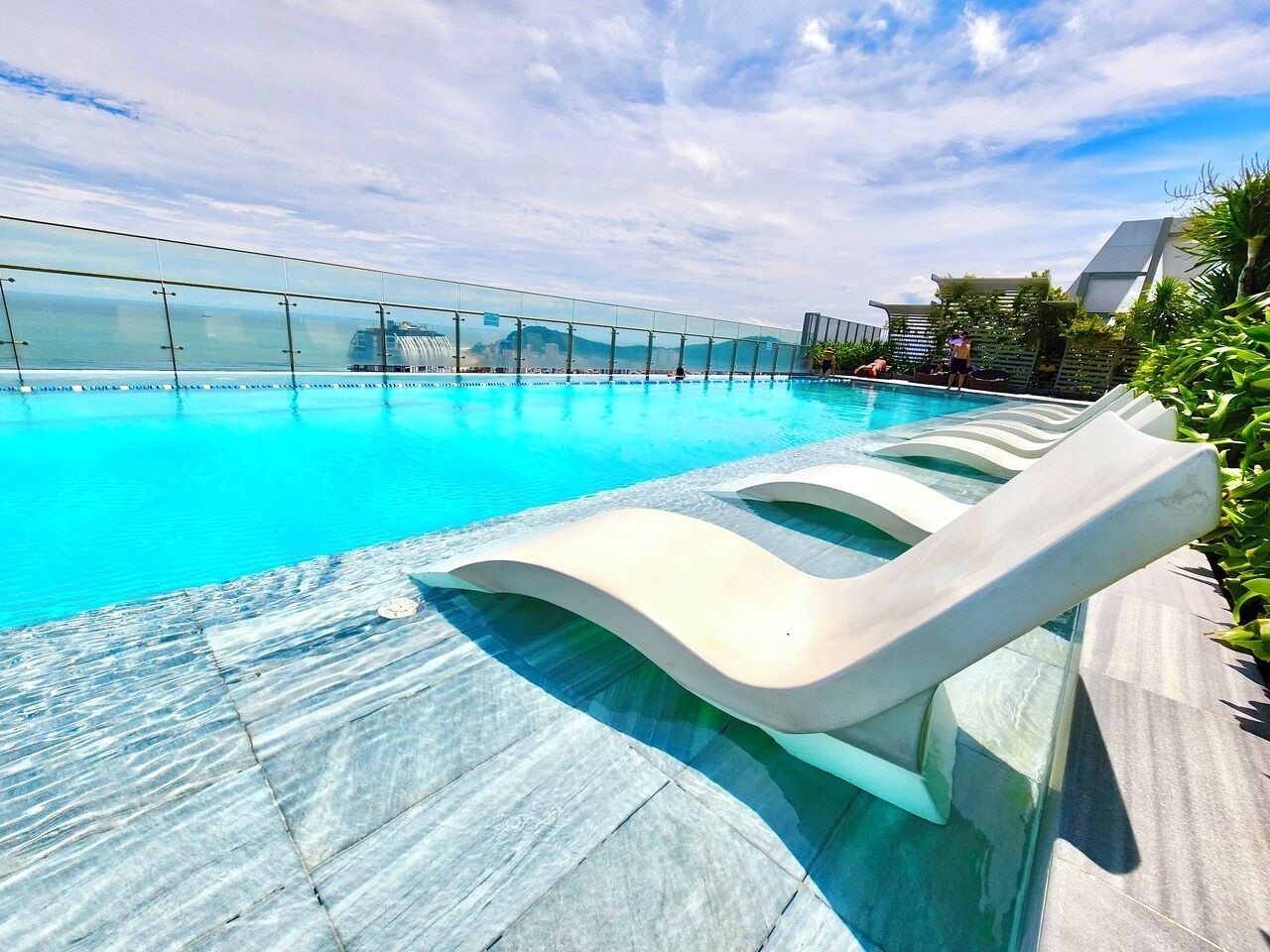 Piscina com barreira de vidro, uma cadeira de descanso branca e vista para o oceano.