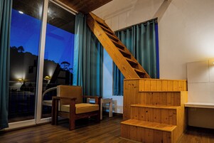 Quarto com escada de madeira, uma poltrona estofada, uma mesa pequena e vista para o exterior através de amplas janelas.