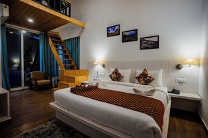 Quarto de hotel com uma cama grande, escada de madeira, mesa e uma cadeira. Há duas luminárias na parede e uma janela com cortinas.